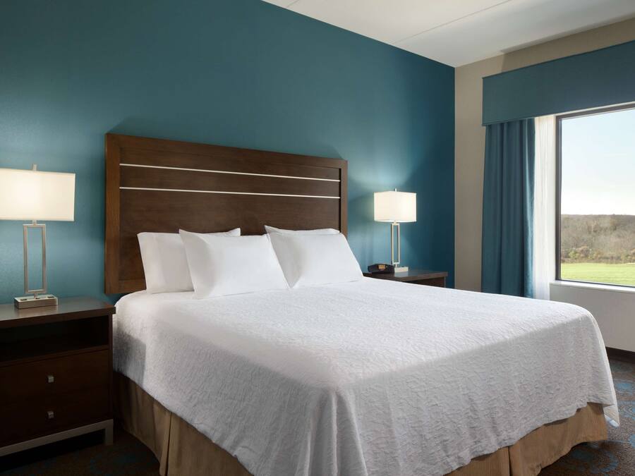 Quarto de hotel com uma cama grande, duas mesas de cabeceira com abajures, vista para o exterior por uma janela e uma parede azul.