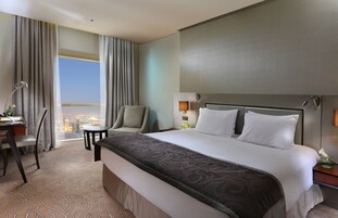 Quarto de hotel com uma cama grande, uma mesa, uma cadeira e vista para a cidade.