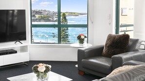Sala moderna com uma janela ampla que oferece vista para a costa, uma TV de tela plana, um sofá de couro e uma mesa de centro com flores.