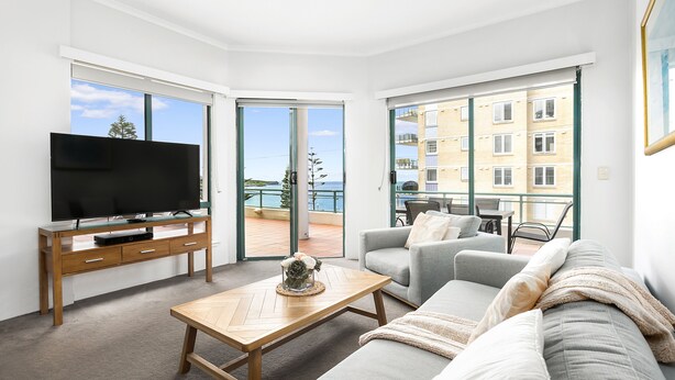 Sala de estar moderna com televisão de tela plana, mesa de centro de madeira, sofá cinza e varanda com vista para o mar.