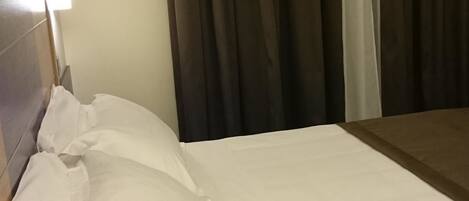 Uma cama bem arrumada com lençóis e almofadas brancos, um abajur e uma janela com cortinas escuras.