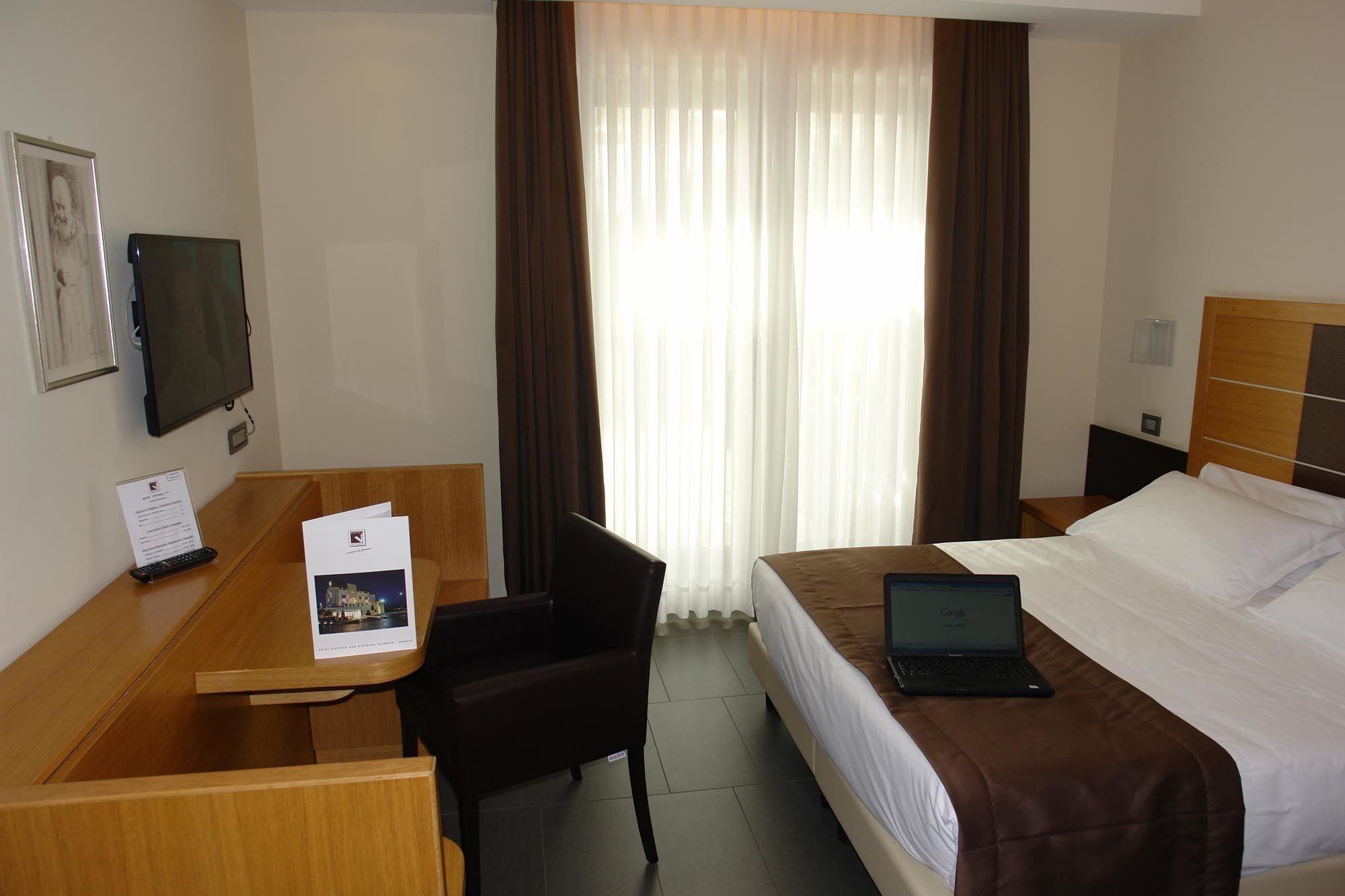 Quarto de hotel com cama, escrivaninha com laptop, cadeira, televis ão e janela com cortinas.