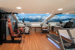 Uma academia com diversos equipamentos de exercício, incluindo esteiras, elípticos e aparelhos de musculação, com uma grande janela que oferece vista para as montanhas.