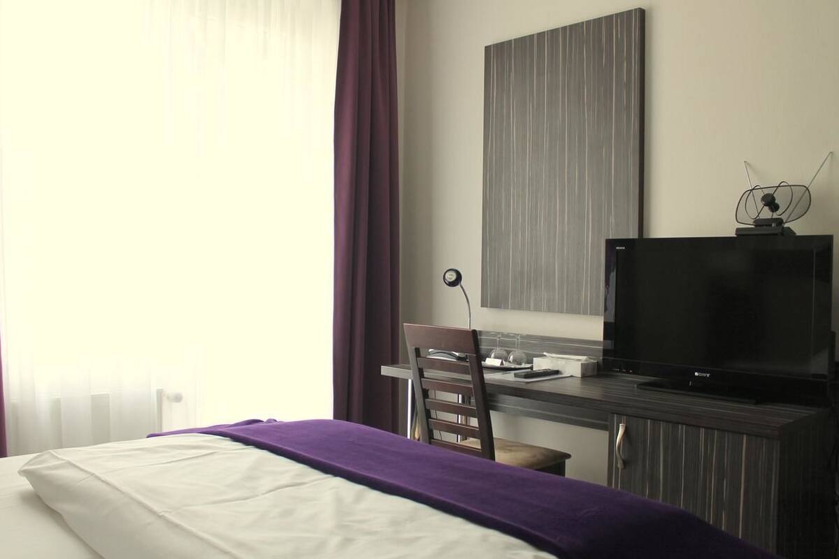 Quarto de hotel com cama, escrivaninha com televisão e janela com cortinas.