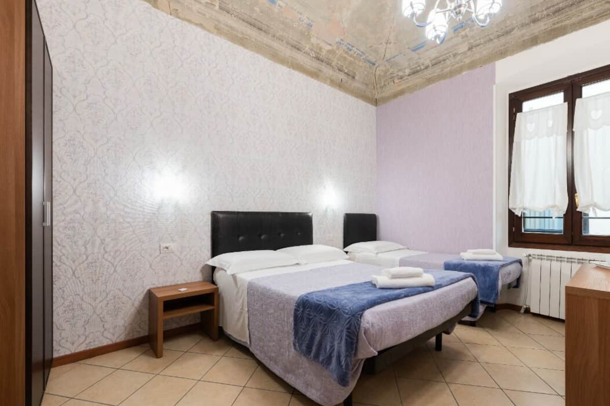 Quarto com duas camas, um banquinho e janela com cortinas.