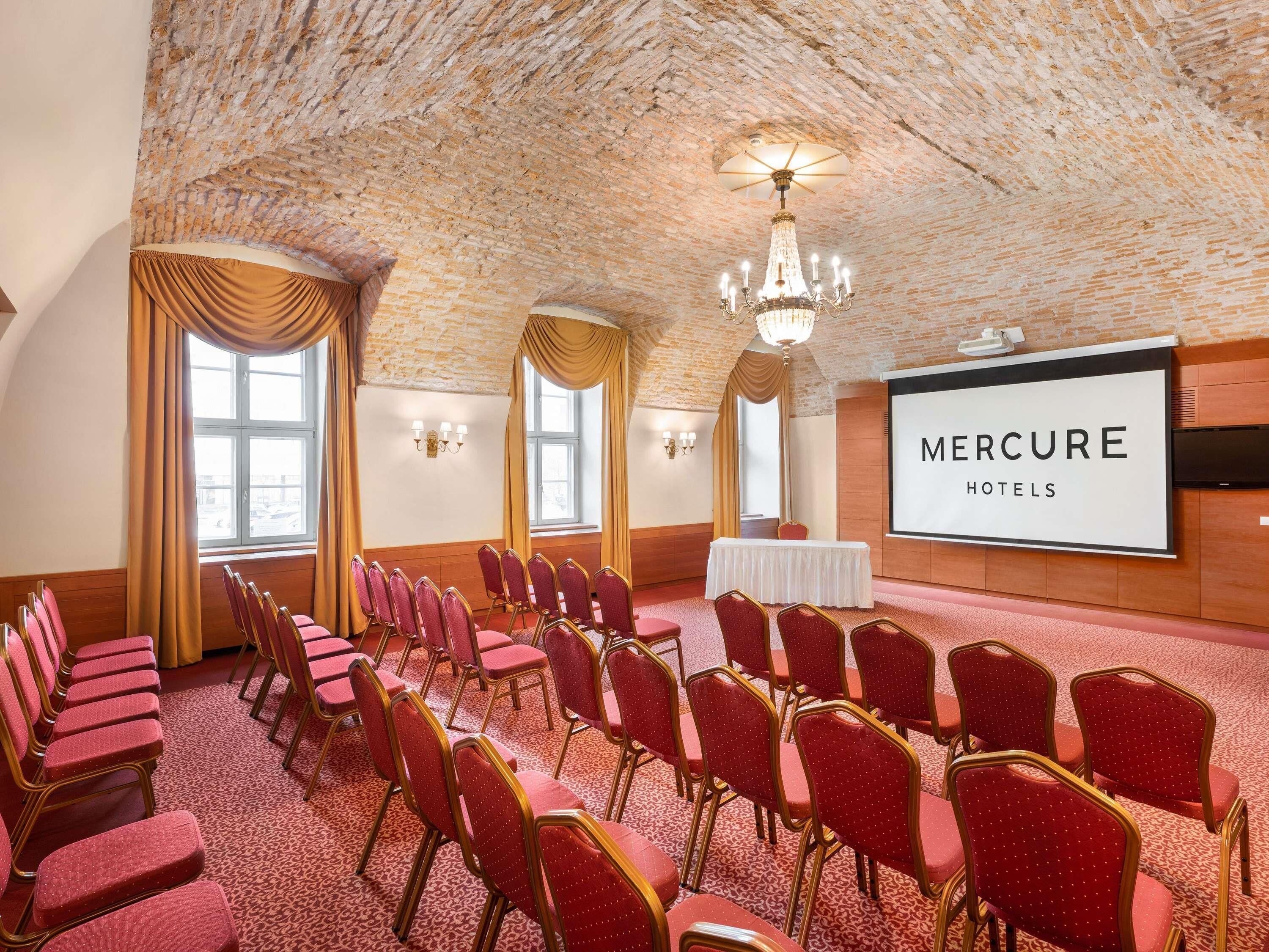 Uma sala de conferências com cadeiras vermelhas, um candelabro e uma tela grande exibindo 'Mercure Hotels'.