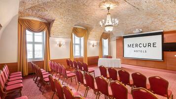 Uma sala de conferências com cadeiras vermelhas, um candelabro e uma tela grande exibindo 'Mercure Hotels'.