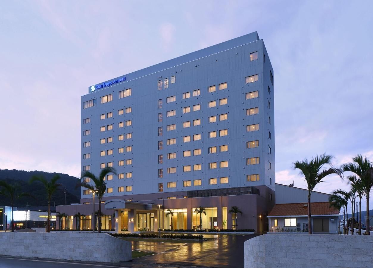 Um hotel com vários andares, fachada plana, diversas janelas e uma placa com a inscrição 'Sun Days Amami'.