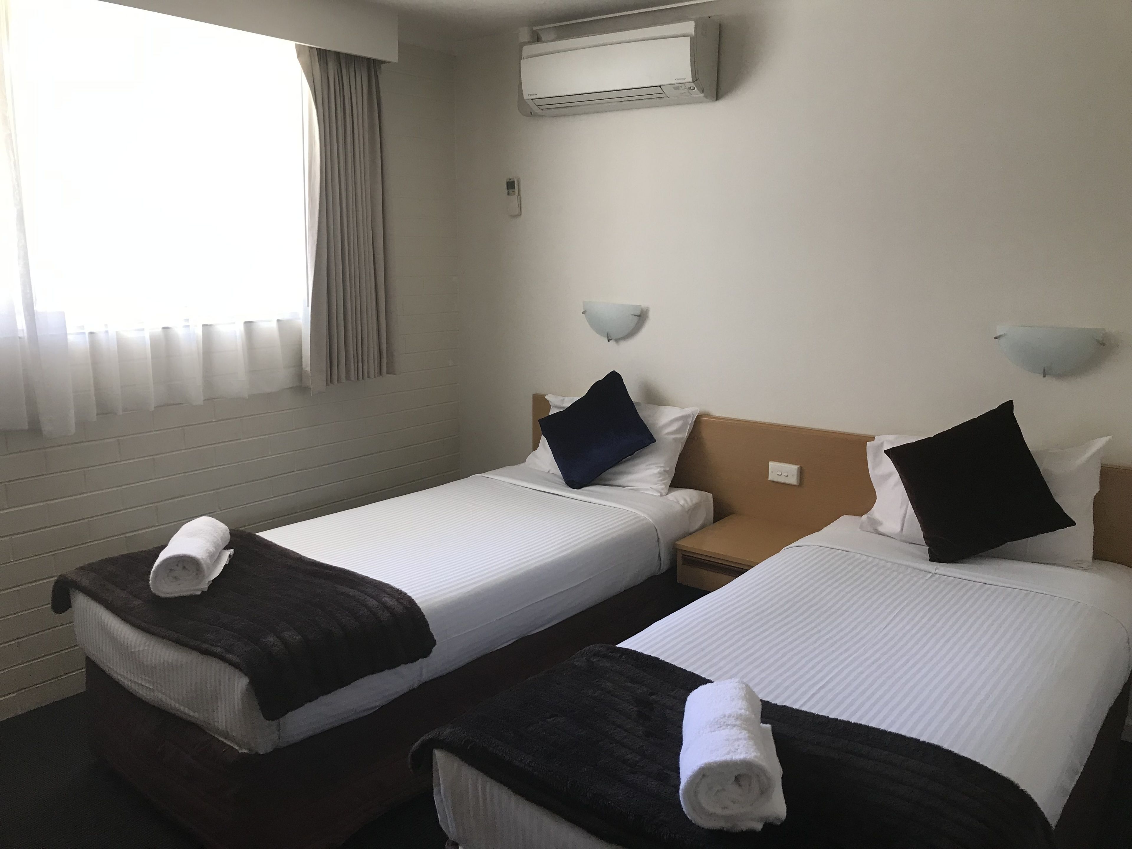 Quarto de hotel com duas camas de solteiro, uma janela com cortinas, unidade de ar condicionado e luminária fixada na parede.
