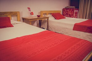 Quarto de hotel com duas camas, cada uma com manta e almofadas vermelhas, cabeceira de madeira, uma mesinha com uma tigela e um livro vermelho na parede.