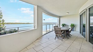 Uma varanda com vista para a água, uma mesa com cadeiras e plantas em vasos.