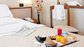 Uma cama bem arrumada com lençóis brancos, uma mesinha de cabeceira com abajur, um vaso com flores secas e uma bandeja de café da manhã com croissants, laranjas e um copo de suco de uva.