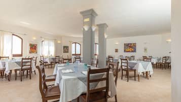 Uma sala de jantar ampla, com várias mesas e cadeiras, janelas grandes e arte decorativa nas paredes.