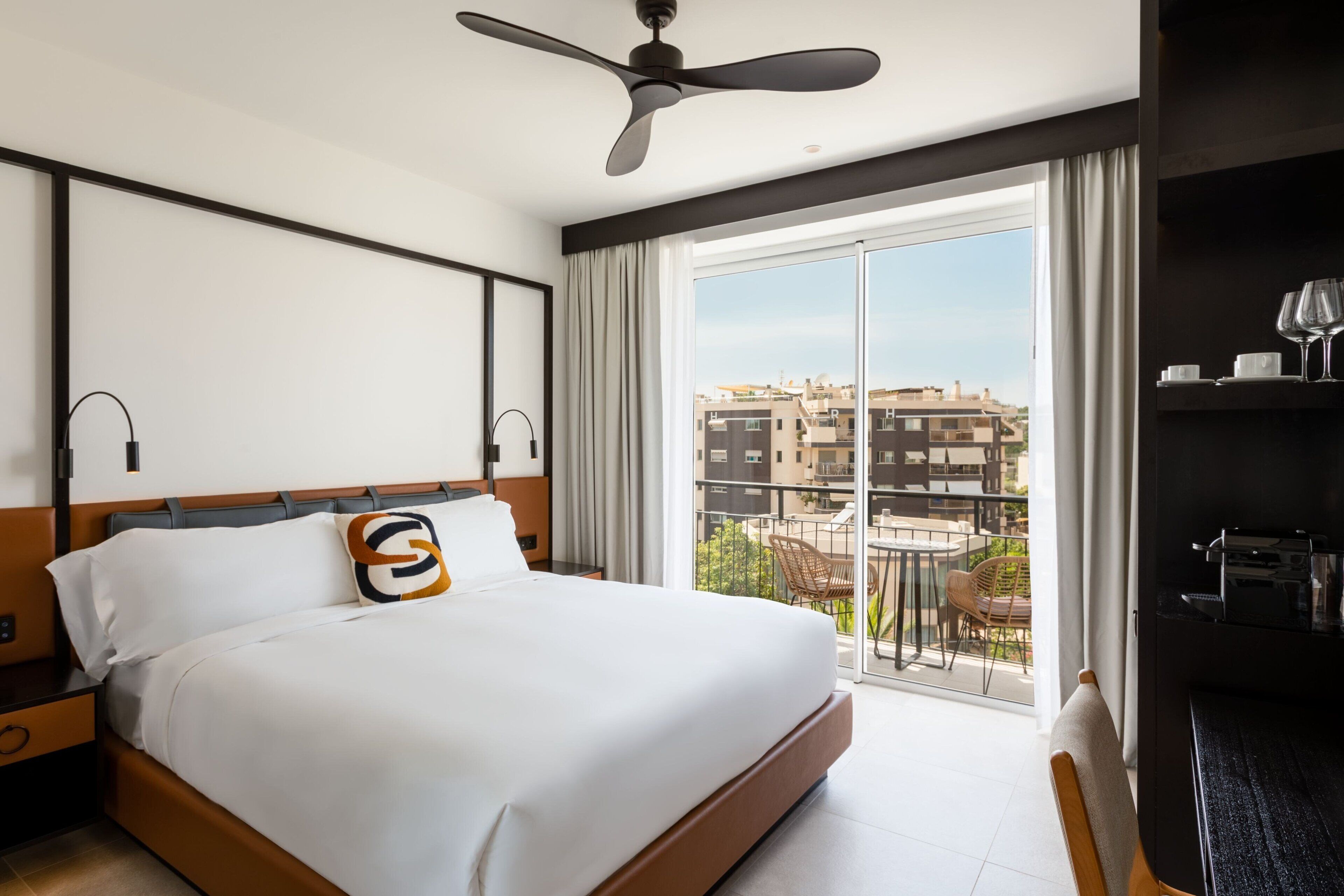 Quarto de hotel com uma cama grande, um ventilador de teto, varanda com vista para edifícios e um espaço de refeições com cadeiras e mesas.