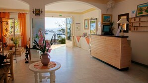 Um saguão de hotel com balcão de recepção, área de estar com mesa e cadeiras, e vista para o mar.