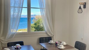 Uma área de refeições com uma mesa posta para quatro pessoas, vista para o mar através de uma janela e uma luminária fixada na parede.