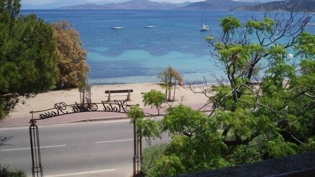 Uma estrada costeira com um banco e árvores, que leva a uma praia com barcos e montanhas ao fundo.