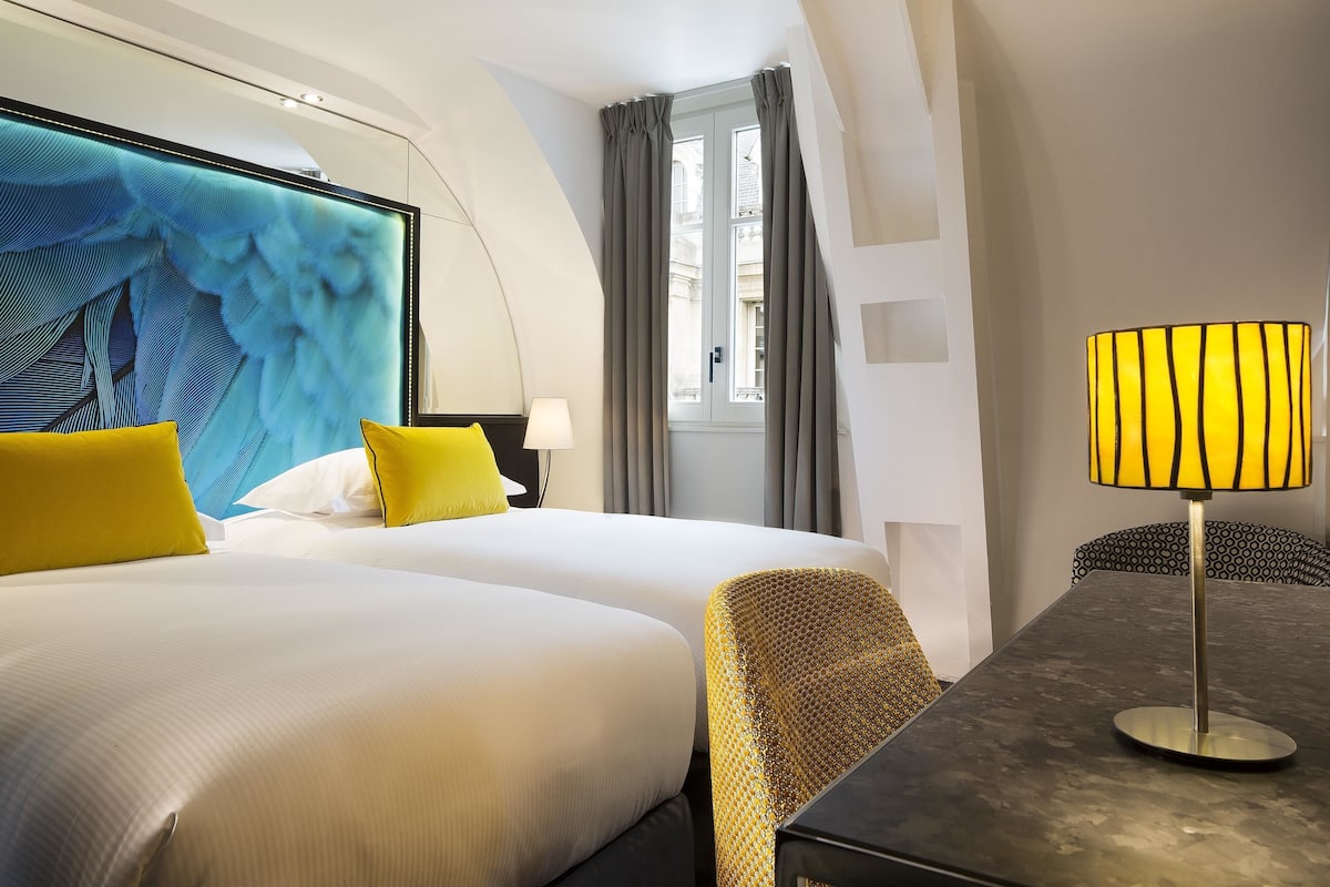 Quarto de hotel com duas camas, uma escrivaninha, uma luminária e uma grande obra de arte abstrata na parede.