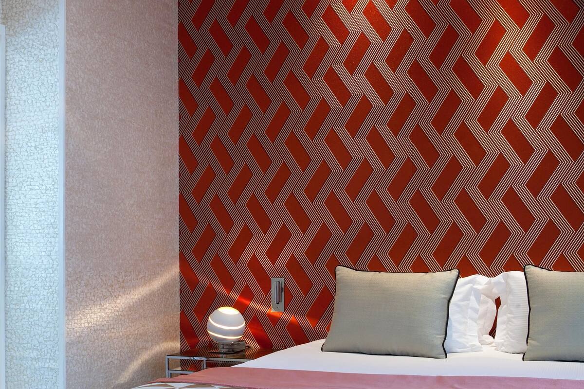 Quarto moderno com parede estampada, cama com colcha de padrão geométrico e luminária de cabeceira.