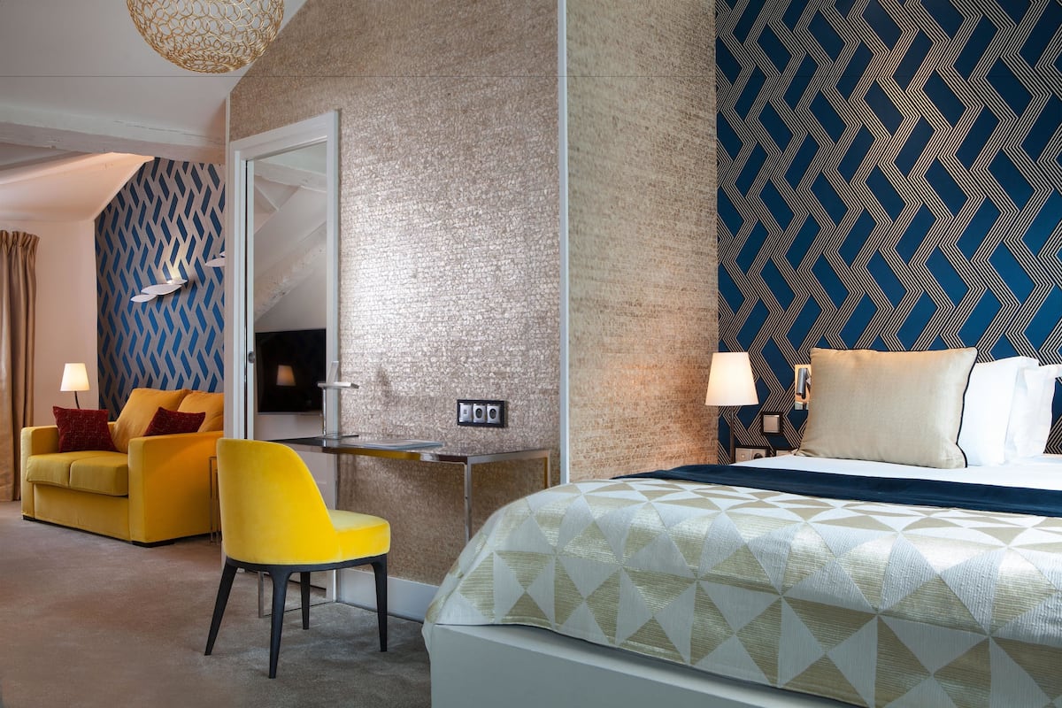 Quarto de hotel com cama, escrivaninha, poltrona amarela e televisão.