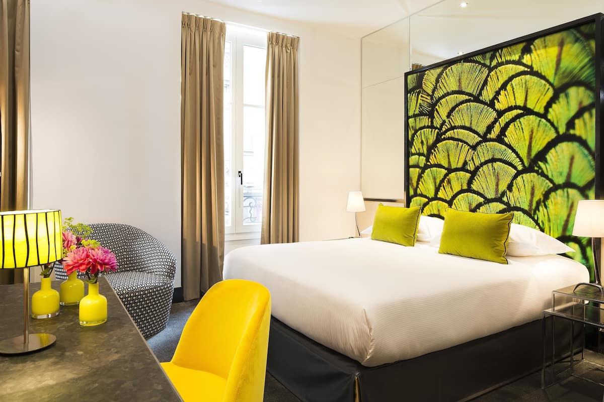 Quarto de hotel com cama, escrivaninha com luminária, cadeira e uma grande obra de arte na parede.