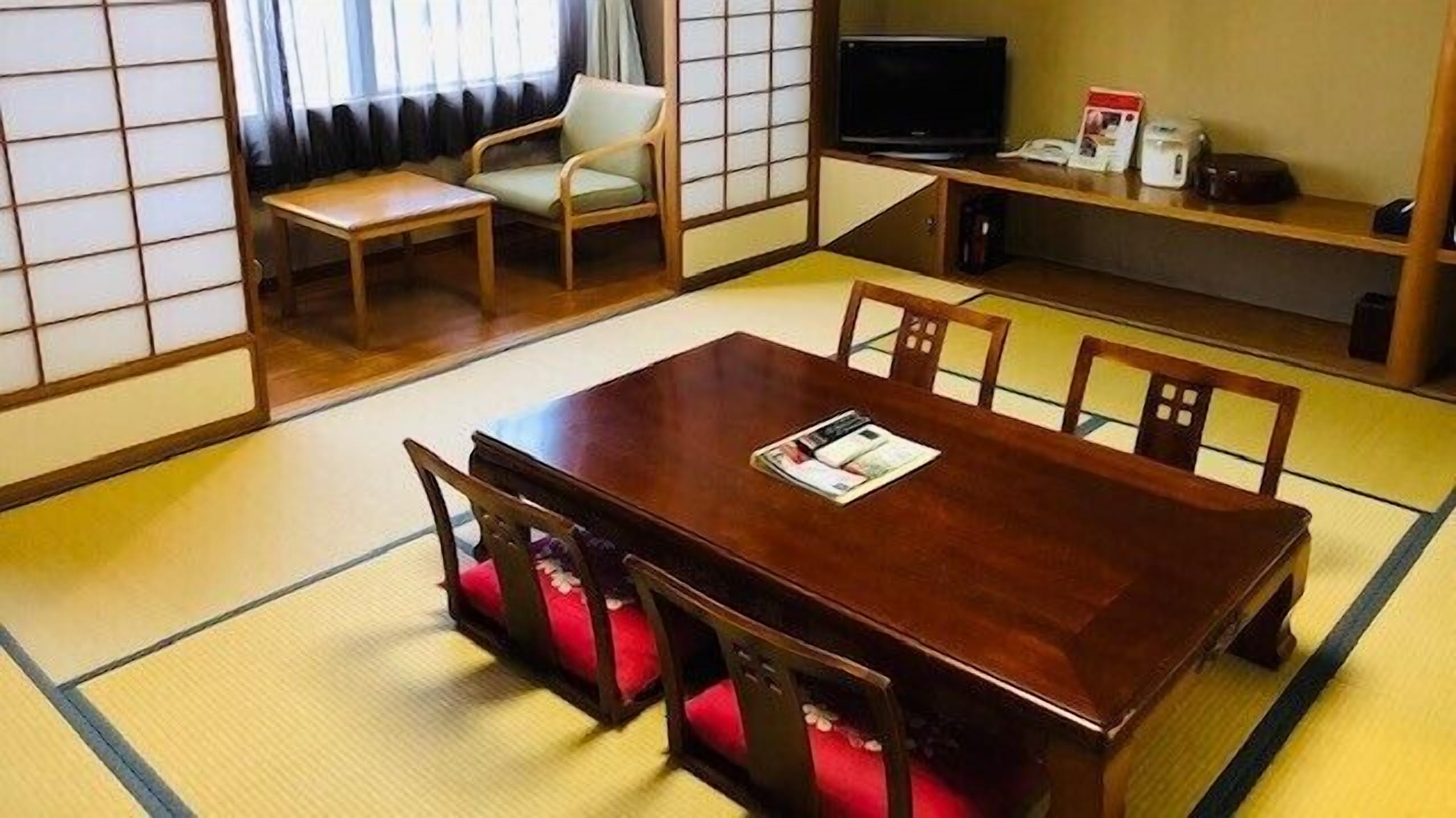Quarto japonês tradicional com tatames, uma mesa baixa com cadeiras, uma televisão e uma estante com objetos.
