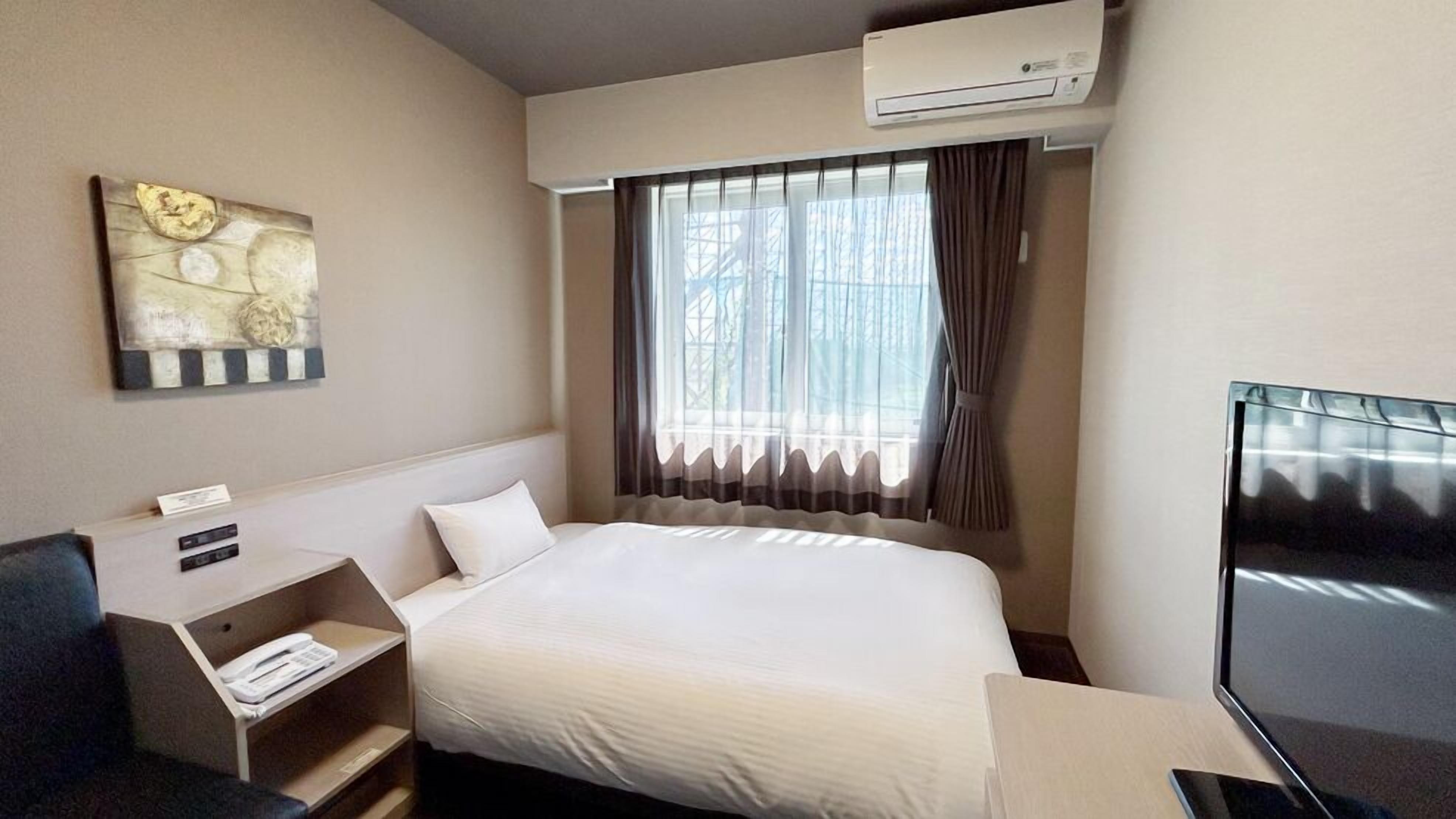 Quarto de hotel com uma cama, uma escrivaninha, uma cadeira, uma televisão e um aparelho de ar condicionado.