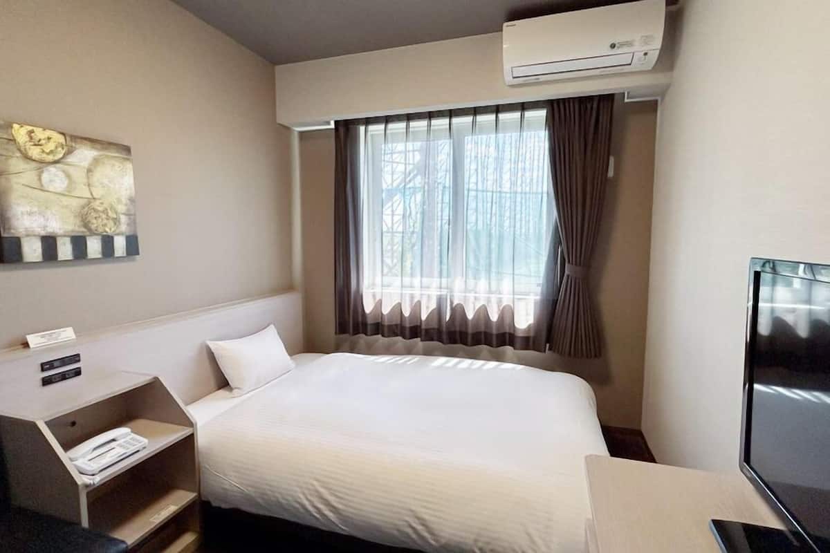 Quarto de hotel com uma cama, uma escrivaninha, uma cadeira, uma televisão e um aparelho de ar condicionado.