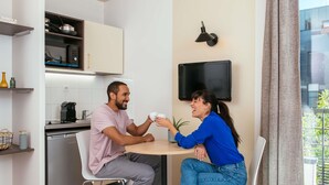 Uma cozinha moderna com geladeira, micro-ondas e cafeteira, uma área de refeições com mesa e cadeiras, e uma TV fixada na parede.
