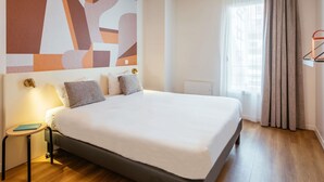 Quarto de hotel com uma cama grande, mesas de cabeceira, uma obra de arte na parede, uma janela com cortinas e um radiador.