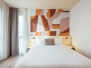 Quarto de hotel com uma cama grande, uma mesa de cabeceira com abajur, uma obra de arte na parede e uma janela com cortinas.