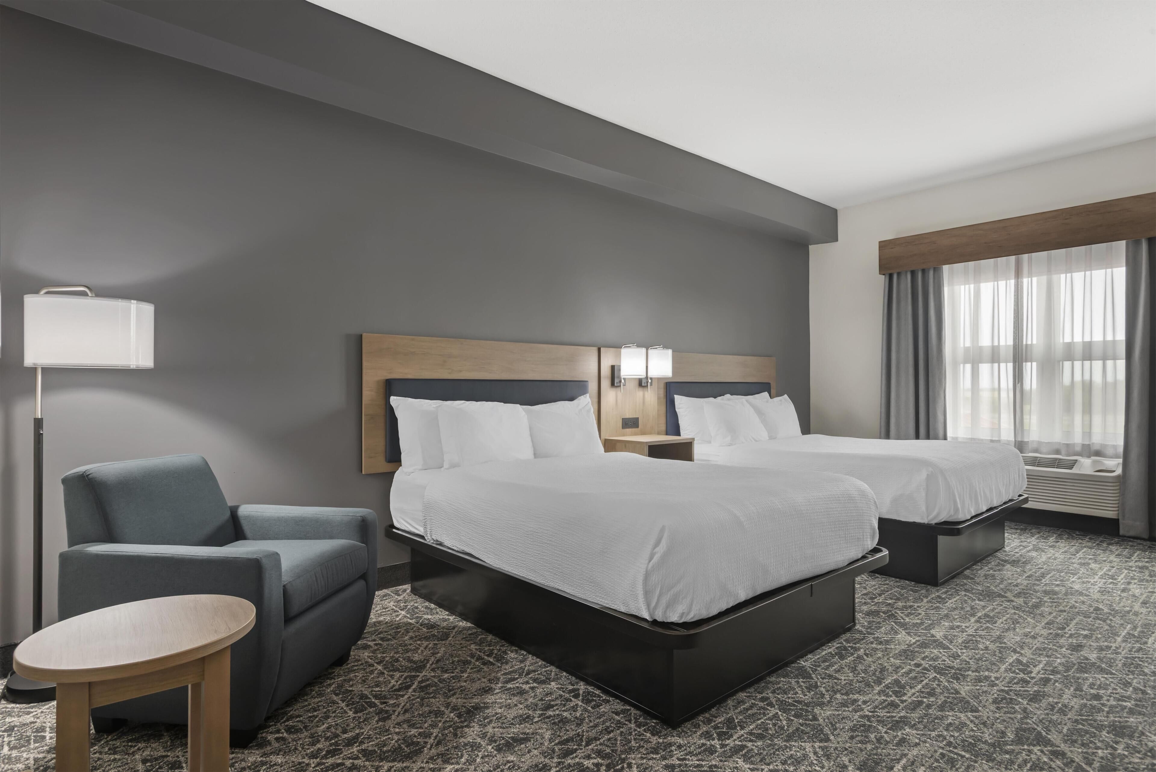 Quarto de hotel com duas camas, uma escrivaninha, uma cadeira, uma luminária e uma janela com cortinas.