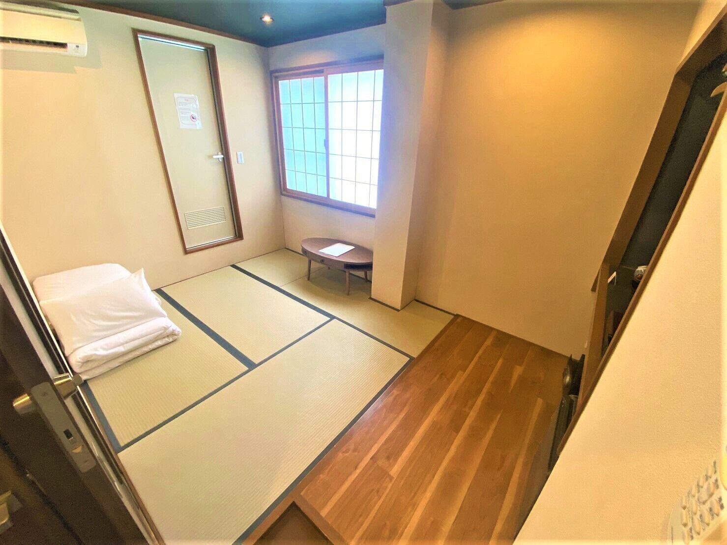 Quarto com tatames, piso de madeira, cama com travesseiro branco, uma pequena mesa e janela com vista.