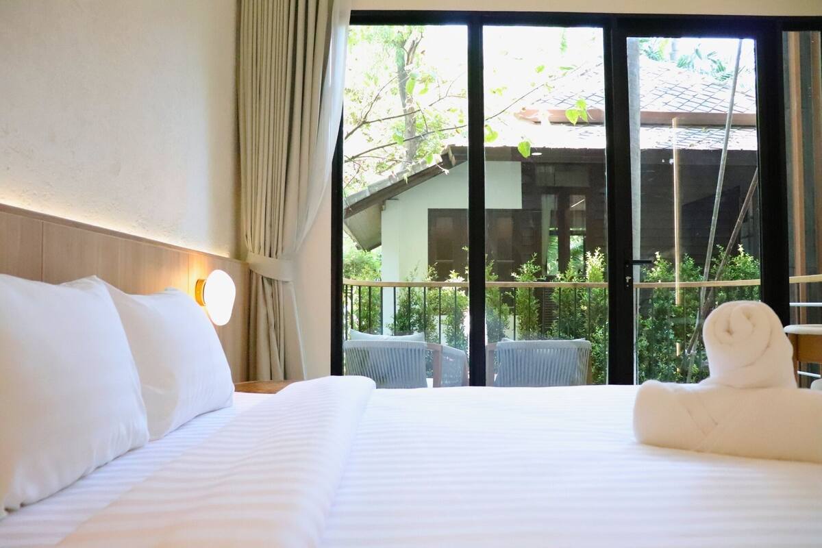 Quarto de hotel com uma cama grande, mesa de cabeceira com abajur e vista para uma varanda com mobiliário exterior.