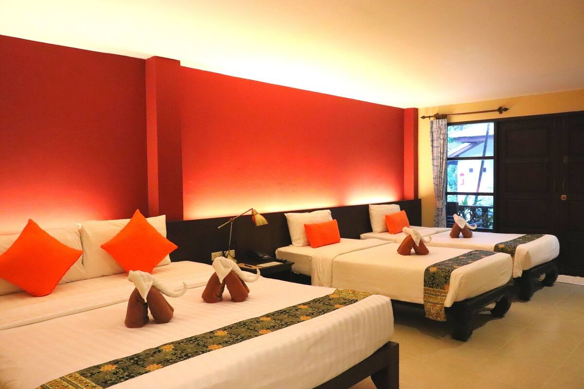 Quarto de hotel com três camas, cada uma com roupa de cama branca e almofadas laranja. Há almofadas decorativas e uma pequena estátua nas camas.