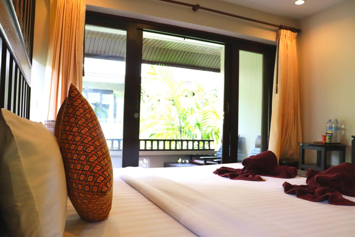 Quarto de hotel com cama, uma janela grande com cortinas, vista para o verde lá fora e uma pequena mesa com garrafas.
