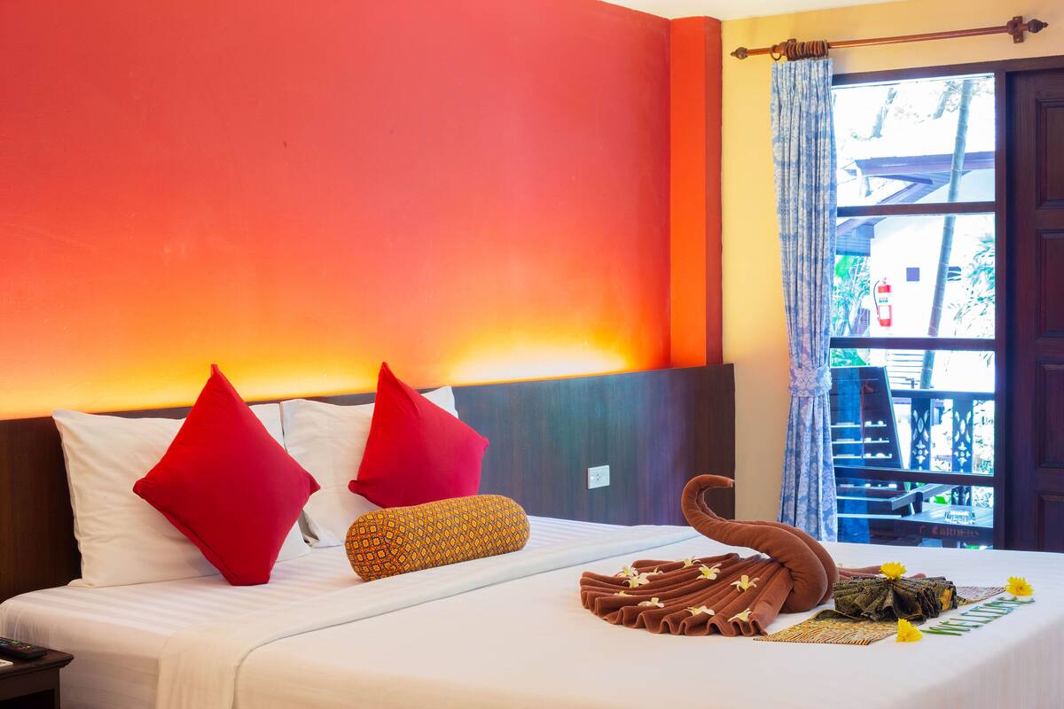 Quarto de hotel com uma cama, almofadas vermelhas estampadas, uma toalha castanha e uma janela com vista para móveis de exterior.