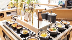 Disposição de buffet com diversos pratos em panelas pretas e cinzentas, um sino de latão e um suporte para cardápio.