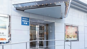 Uma entrada de hotel moderna com fachada de vidro, uma placa azul indicando o quarto número 5 e uma escadaria que leva à entrada.