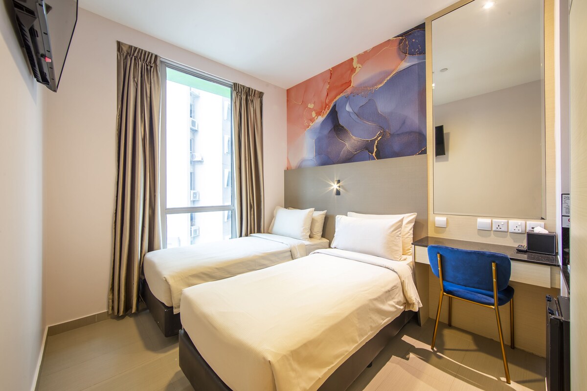 Quarto de hotel com duas camas, uma escrivaninha com cadeira, um espelho grande e uma janela com cortinas.