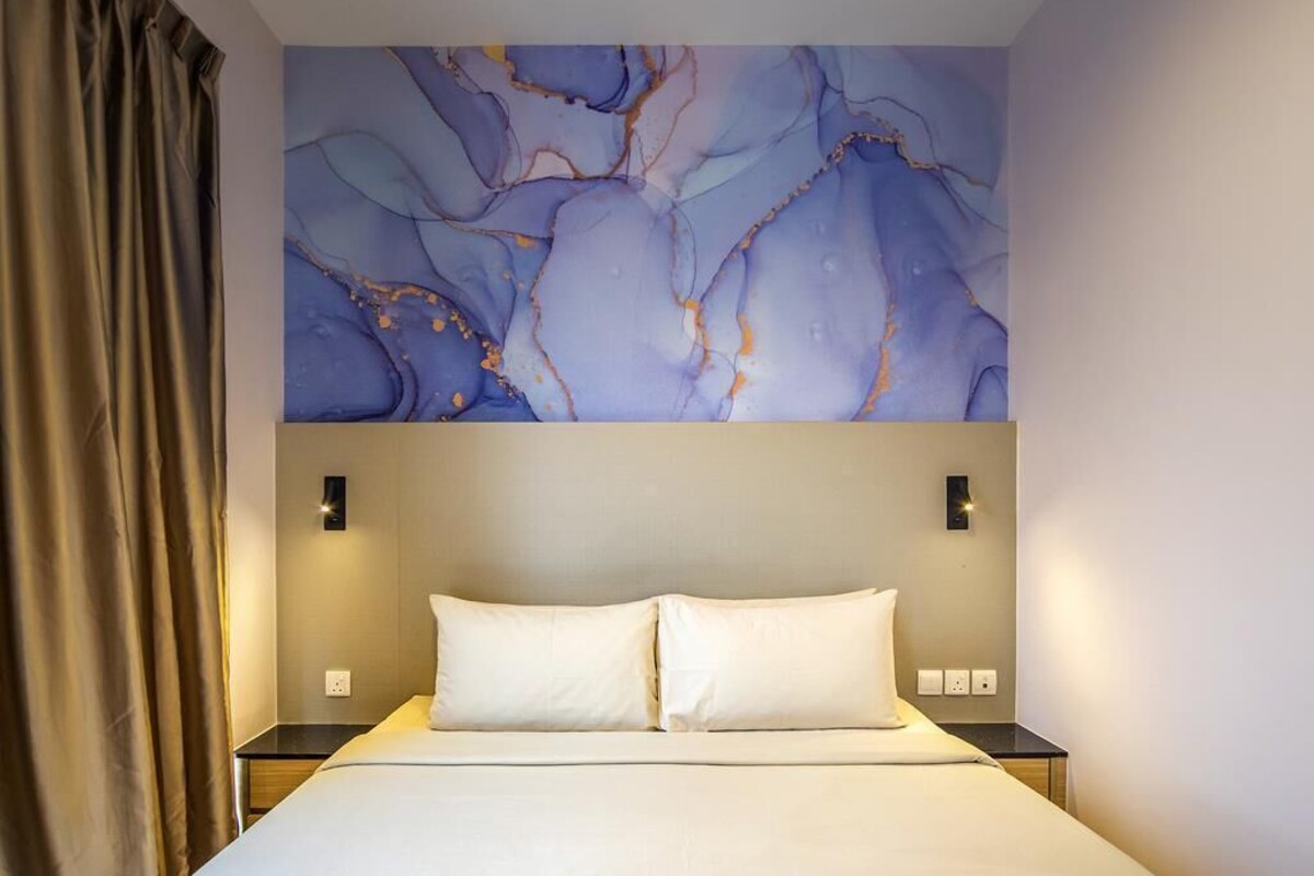 Quarto de hotel com uma cama grande, duas mesas de cabeceira, uma luminária fixada na parede e um quadro decorativo abstrato acima da cama.