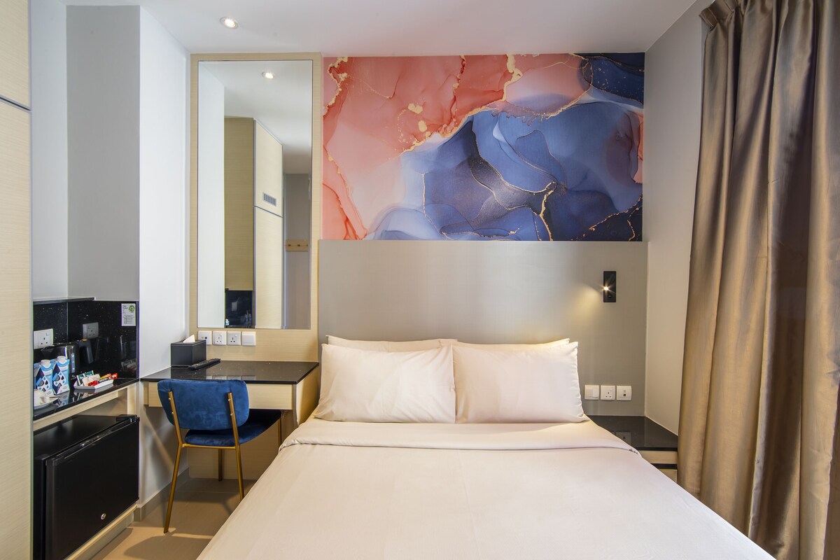Quarto de hotel com cama, escrivaninha e cadeira, pequena cozinha e uma grande pintura abstrata na parede.