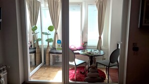 Quarto acolhedor com piso de madeira, uma pequena mesa redonda com cadeiras, uma janela com cortinas e um tapete vermelho.