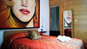 Quarto de hotel com uma cama grande, um edredom vermelho, um travesseiro com estampa de leopardo e um espelho refletindo o rosto de uma mulher.