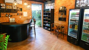Uma área de bar com balcão, banquetes e prateleiras com garrafas e petiscos. Há duas geladeiras repletas de bebidas.