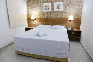 Cama bem arrumada com roupa de cama branca e uma toalha enrolada, duas luminárias de cabeceira e uma cabeceira de madeira.