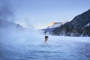 Uma pessoa em uma fonte termal, rodeada por montanhas e árvores cobertas de neve.