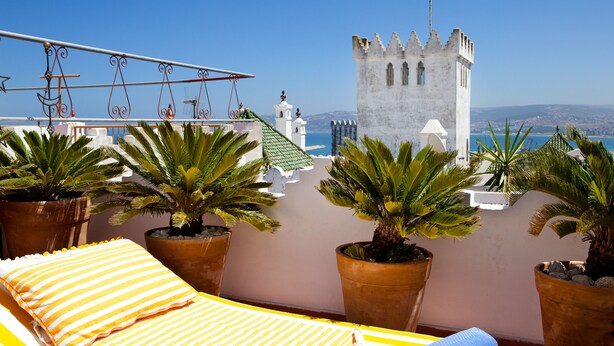 Varanda com cama, plantas em vasos e vista para um castelo e o mar.