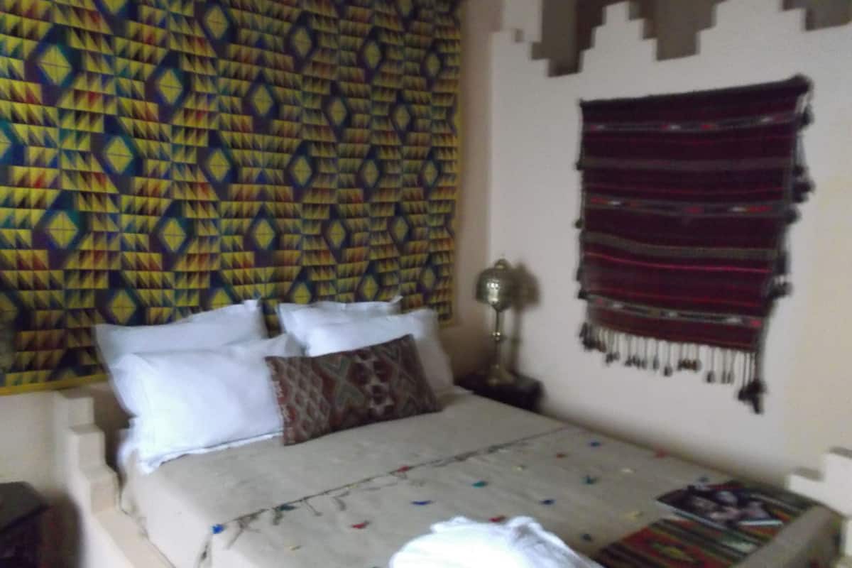 Uma cama com almofadas brancas e estampadas, cabeceira estampada e uma luminária fixada na parede.