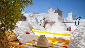 Cama com roupa de cama listrada e chapéu de palha, com vista para um horizonte urbano com edifícios e céu claro.